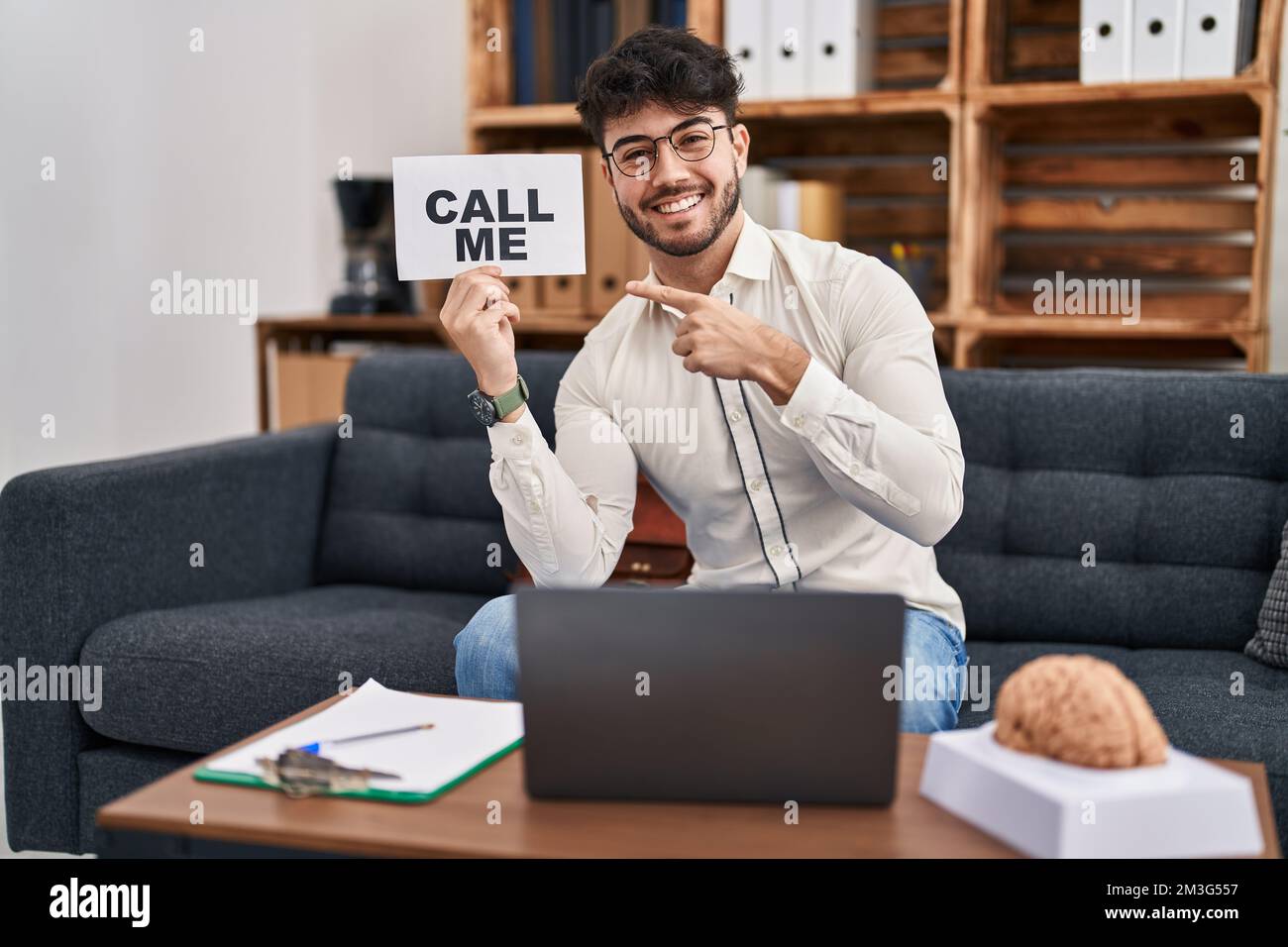 Hispanic man with beard working at therapy office holding call me sign ...