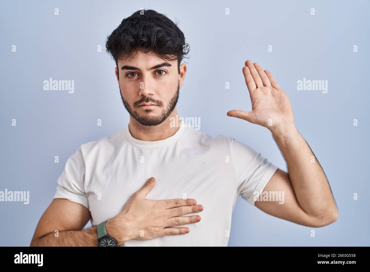 Hispanic man with beard standing over white background swearing with ...