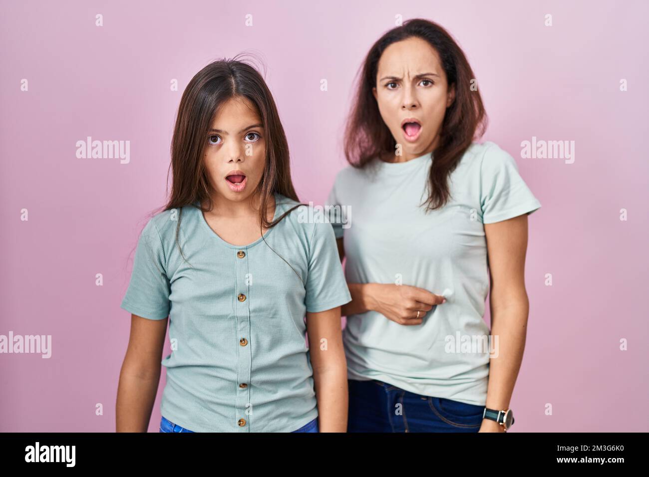 Young mother and daughter standing over pink background in shock face ...