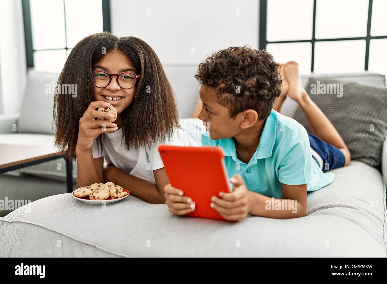 Brother and sister eating cookies using touchpad lying on sofa at home Stock Photo - Alamy