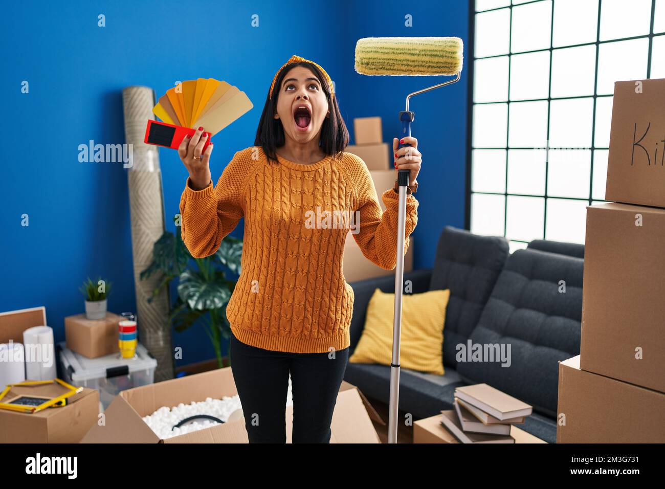 Young hispanic woman painting home walls with paint roller angry and ...