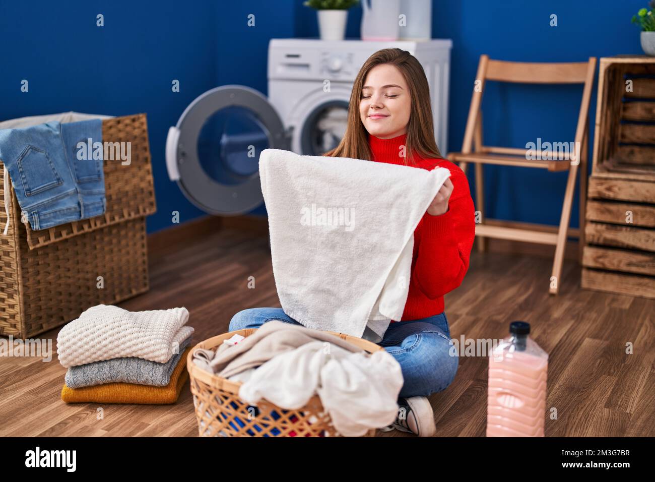 Young blonde woman smelling clothes sitting on floor at laundry room ...