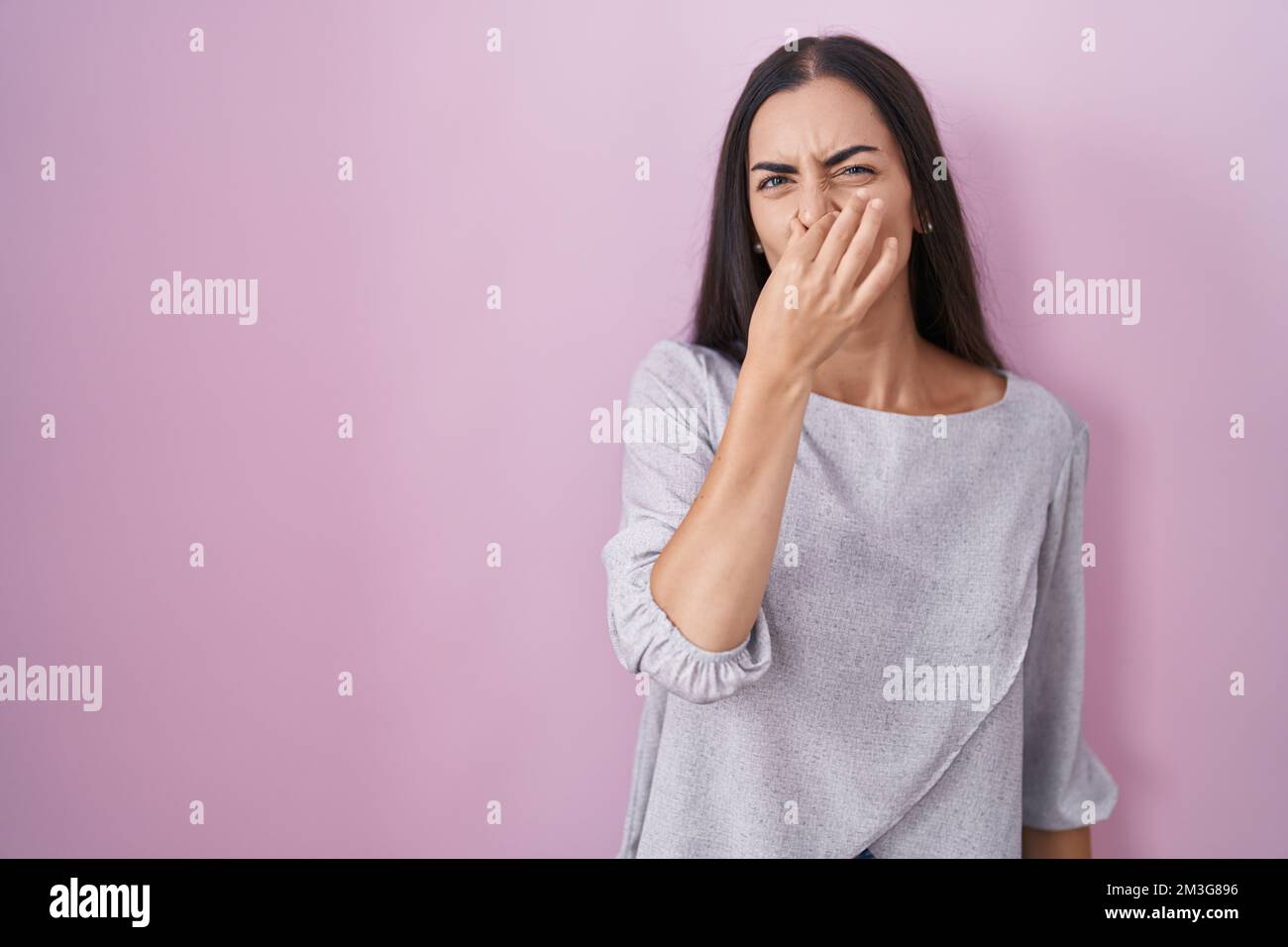 Young brunette woman standing over pink background smelling something ...