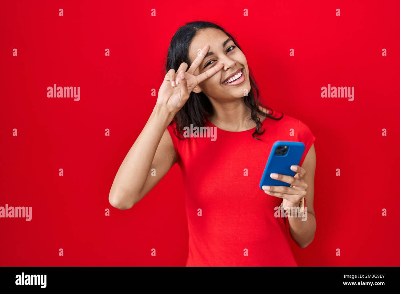 Young brazilian woman using smartphone over red background doing peace ...