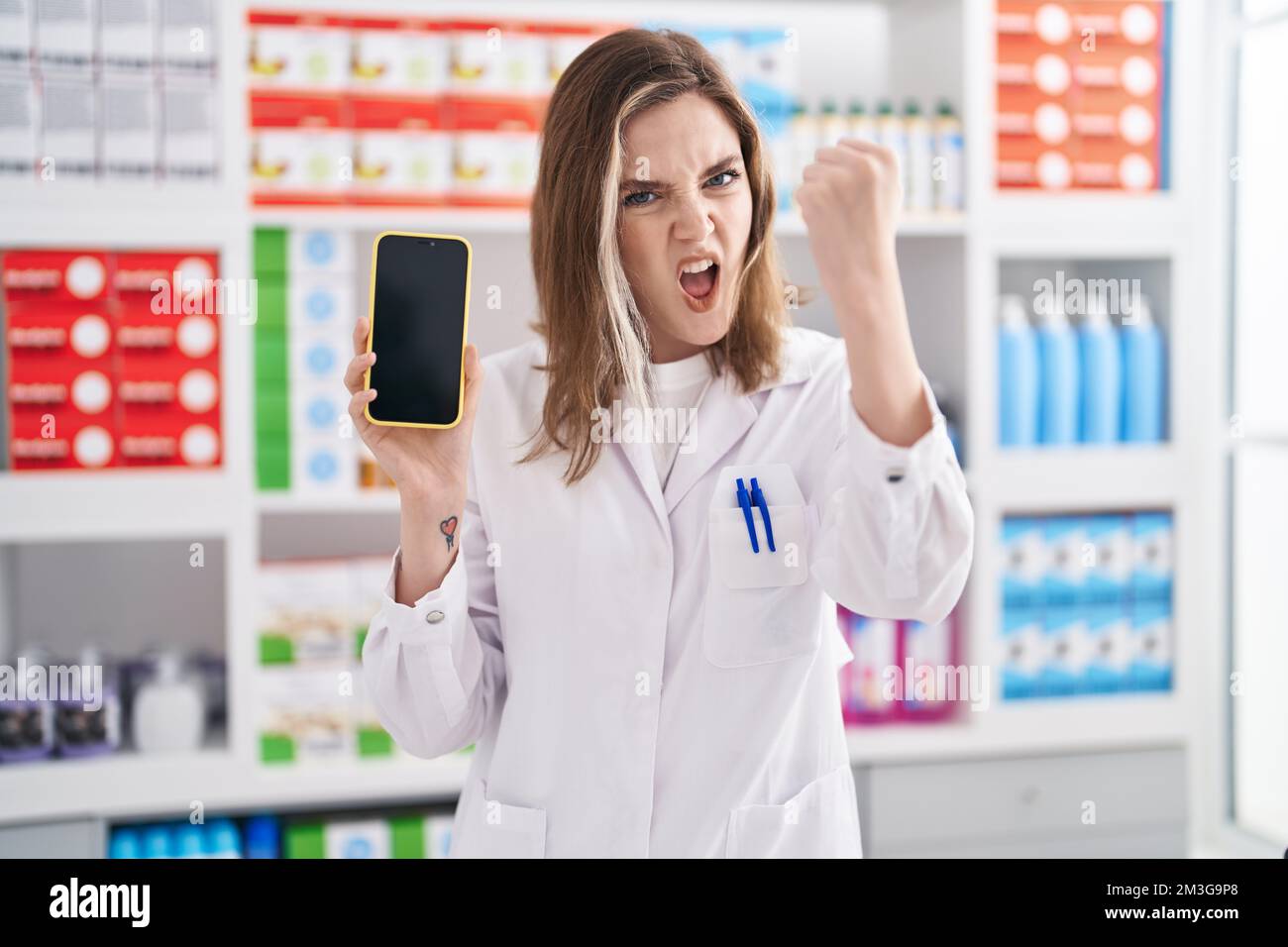 Blonde caucasian woman working at pharmacy drugstore showing smartphone ...