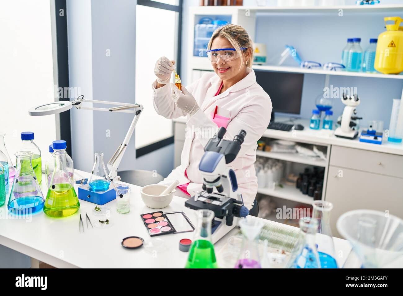 Young hispanic woman wearing scientist uniform holding liquid bottle at ...