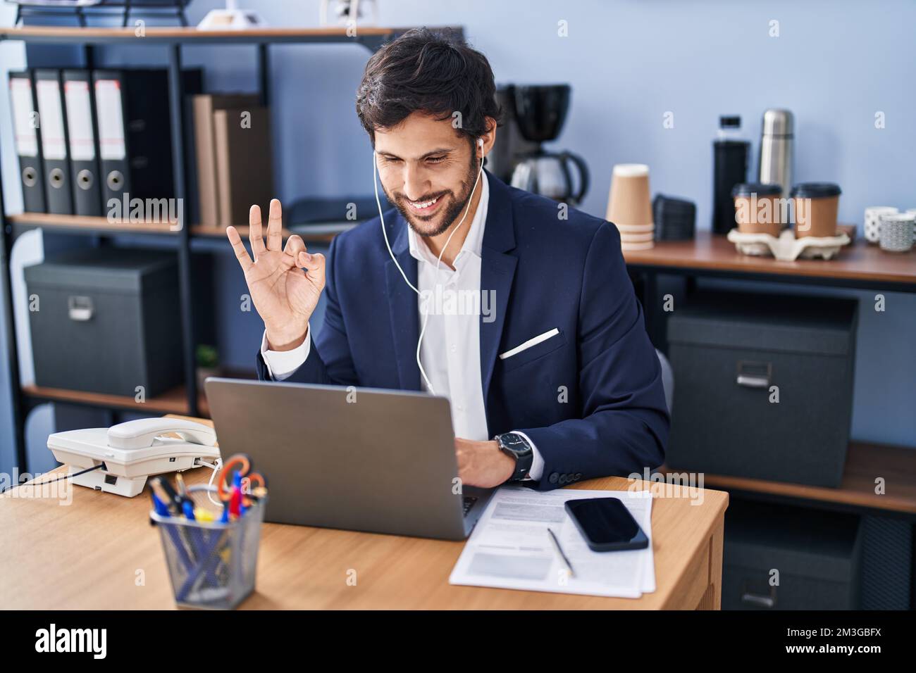 Handsome latin man working at the office using laptop doing ok sign ...