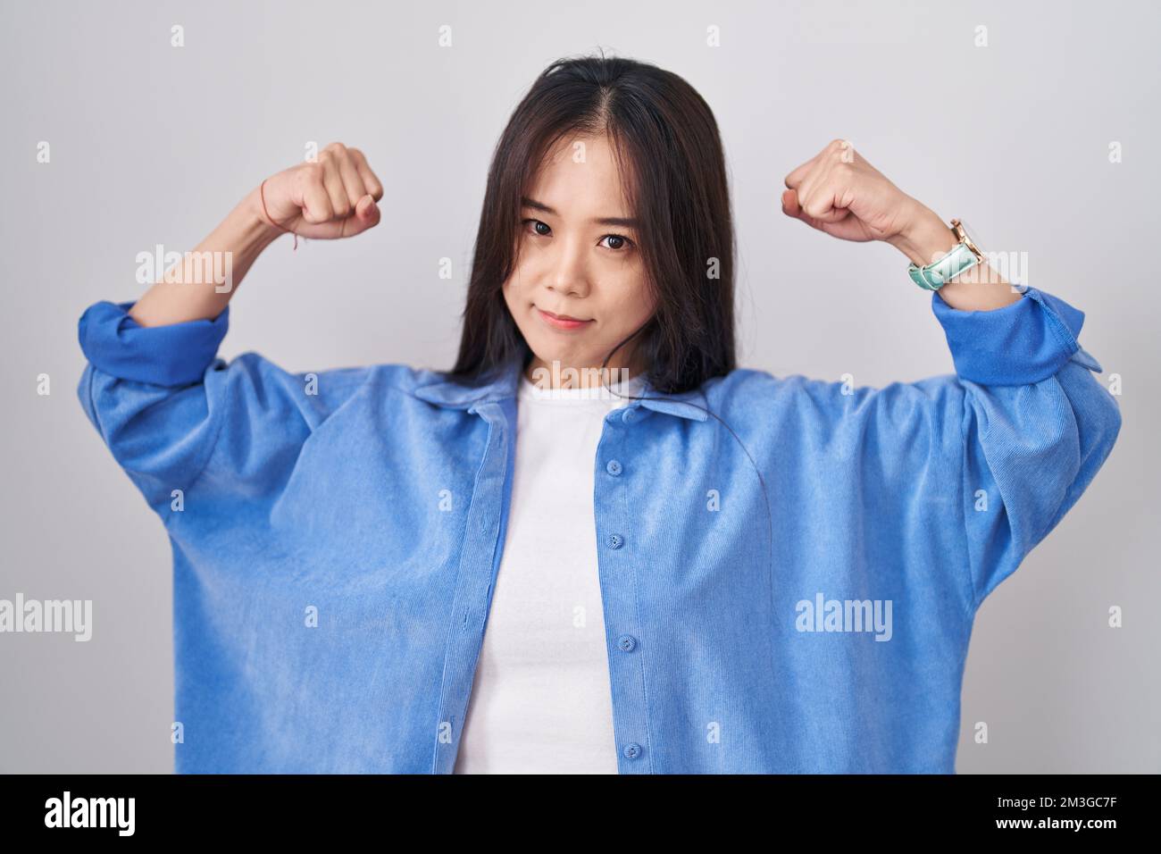 Young chinese woman standing over white background showing arms muscles ...