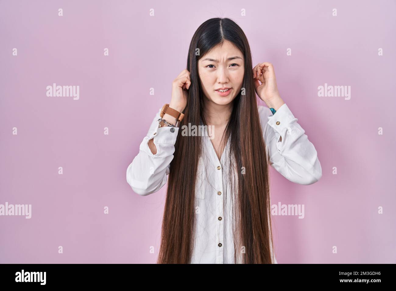 Chinese young woman standing over pink background covering ears with ...