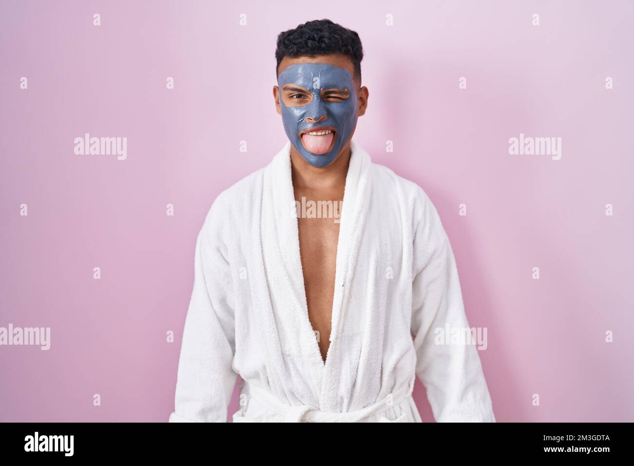 Young hispanic man wearing beauty face mask and bath robe sticking ...