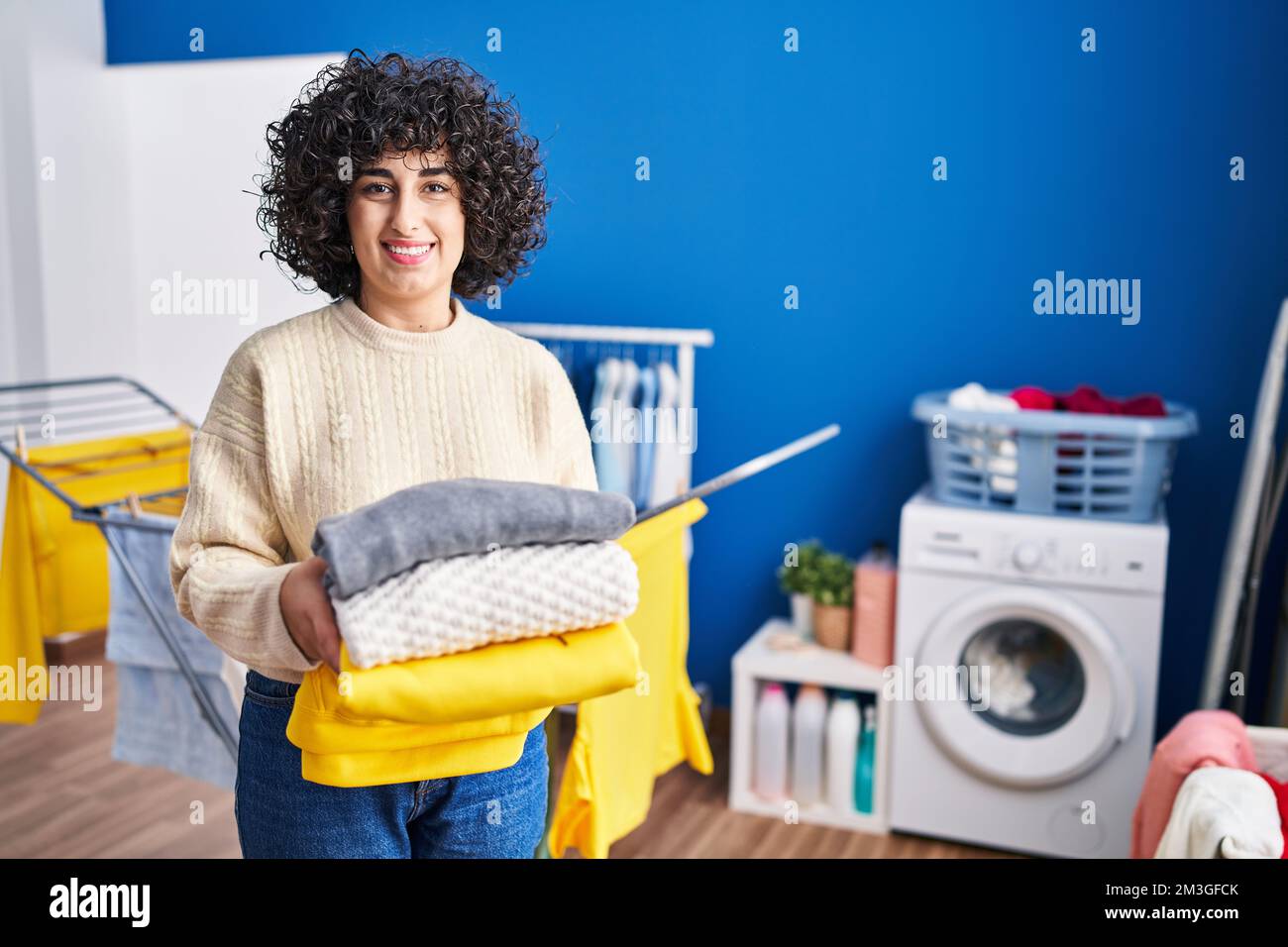 Young brunette woman with curly hair holding clean laundry smiling with a happy and cool smile ...