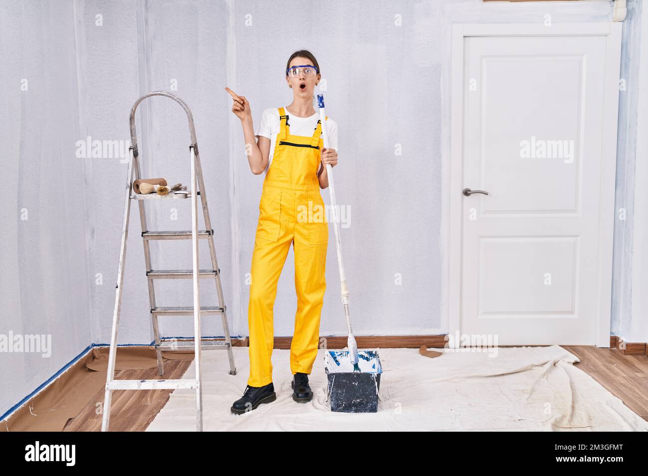 Young caucasian woman painting walls surprised pointing with finger to ...