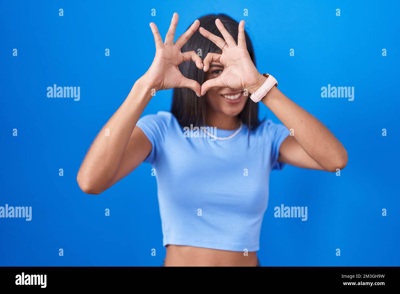 Brunette young woman standing over blue background doing heart shape ...