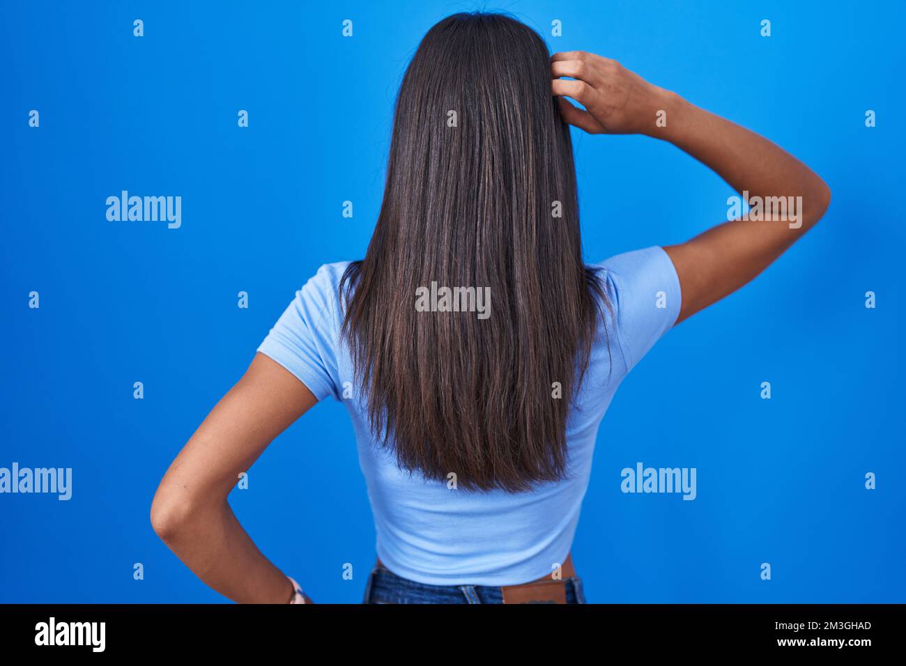 Brunette young woman standing over blue background backwards thinking ...