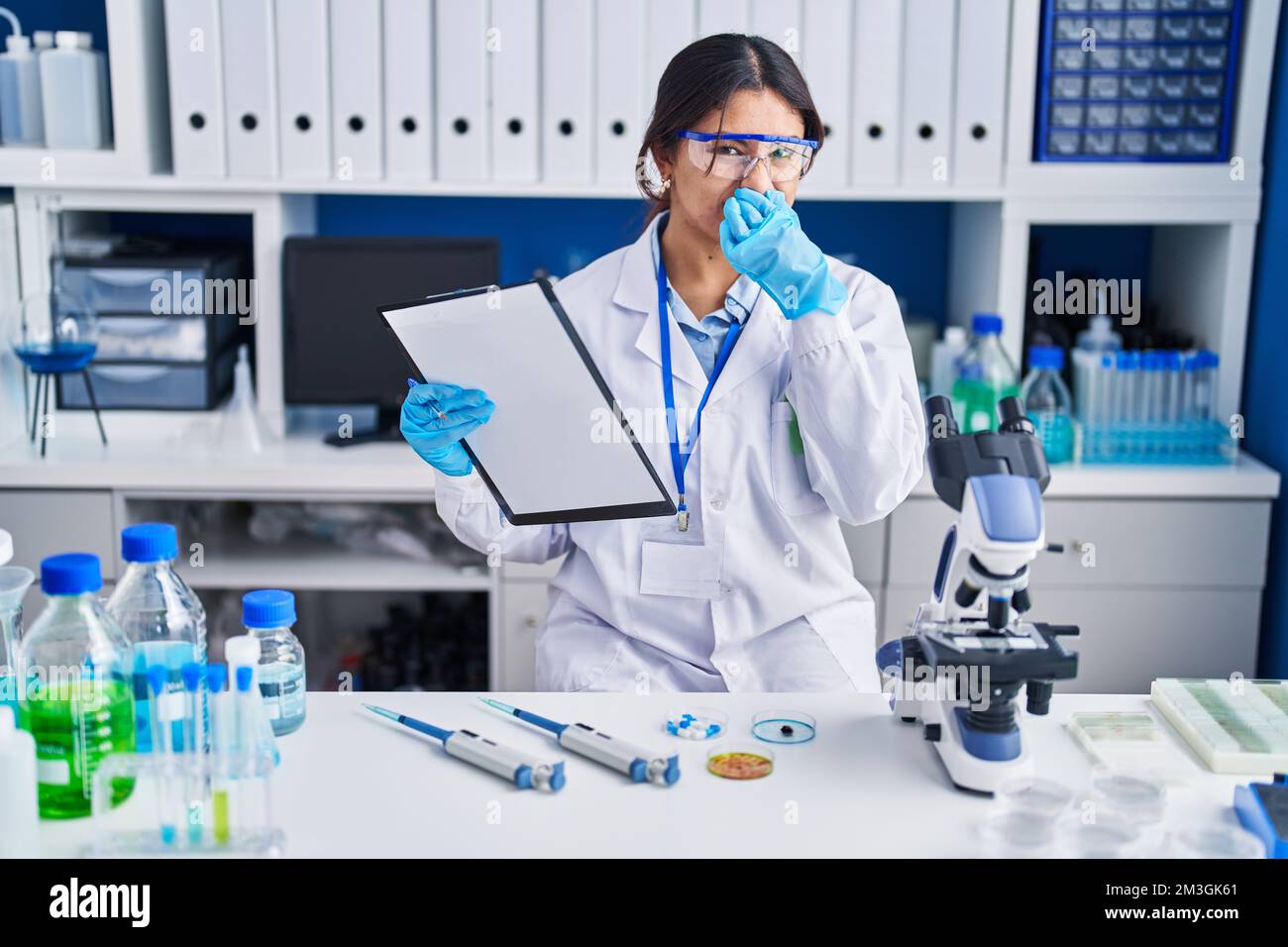 Hispanic young woman working at scientist laboratory smelling something ...