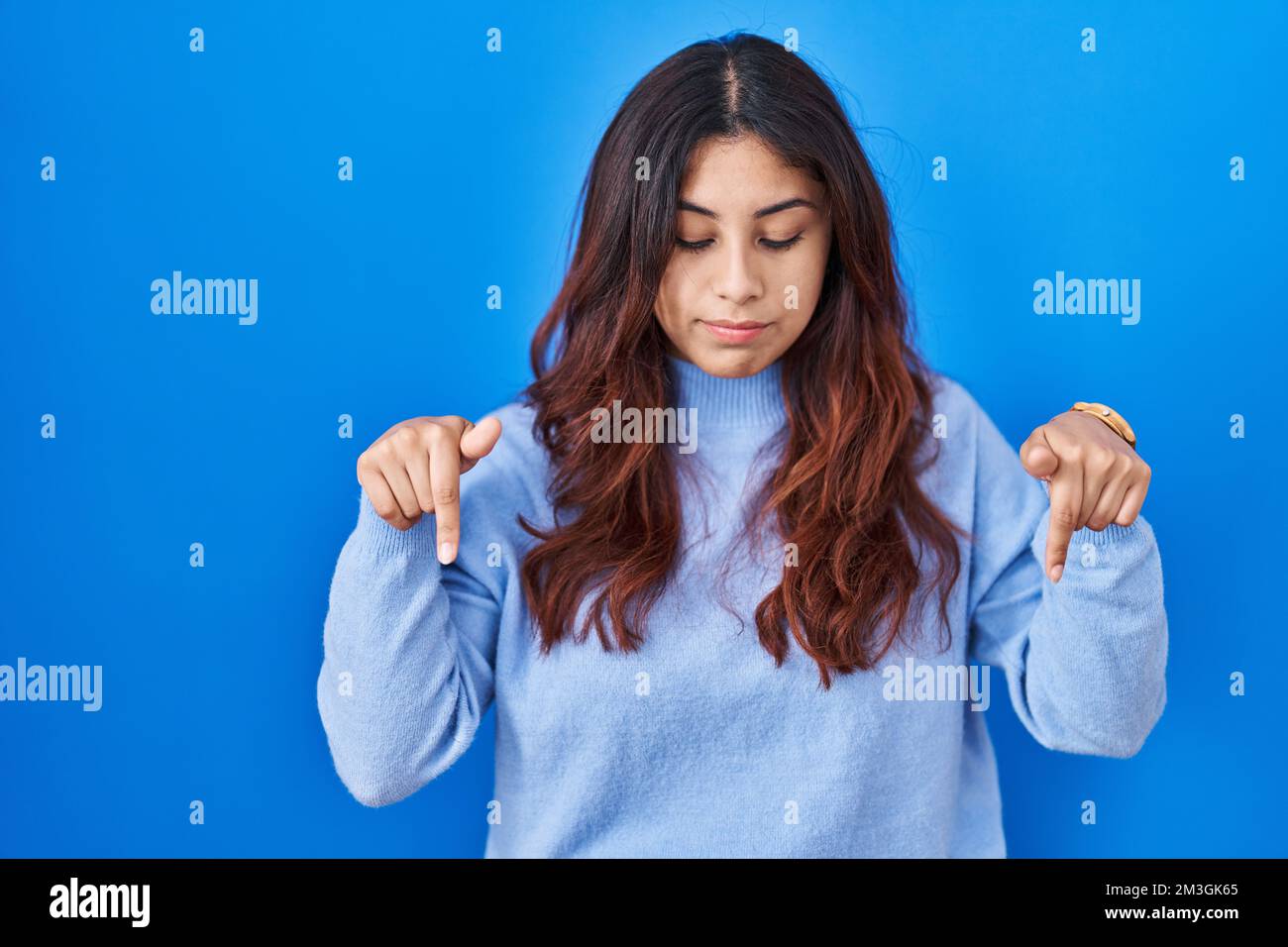 Hispanic young woman standing over blue background pointing down ...