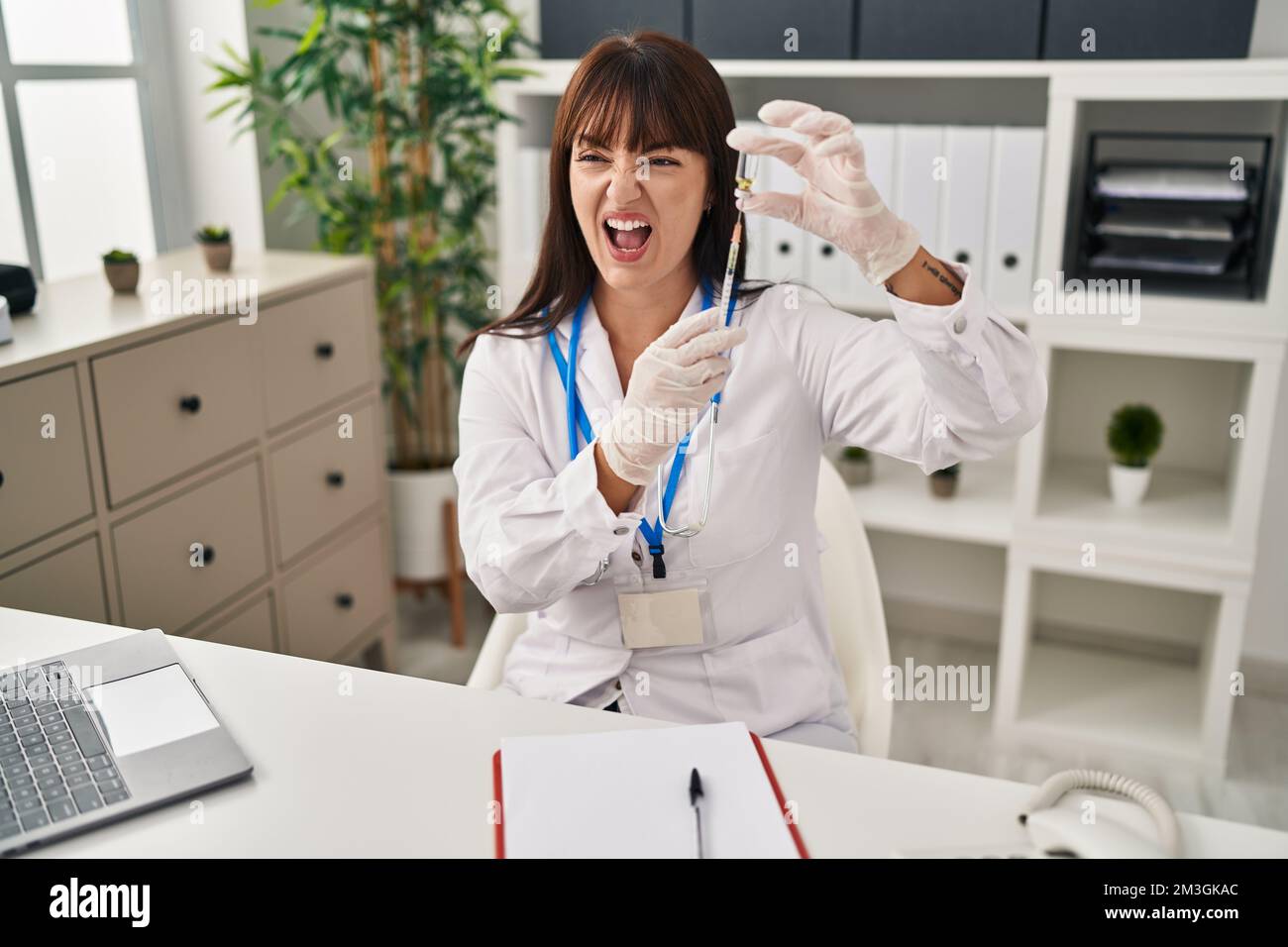Young brunette doctor woman holding syringe and vaccine angry and mad ...