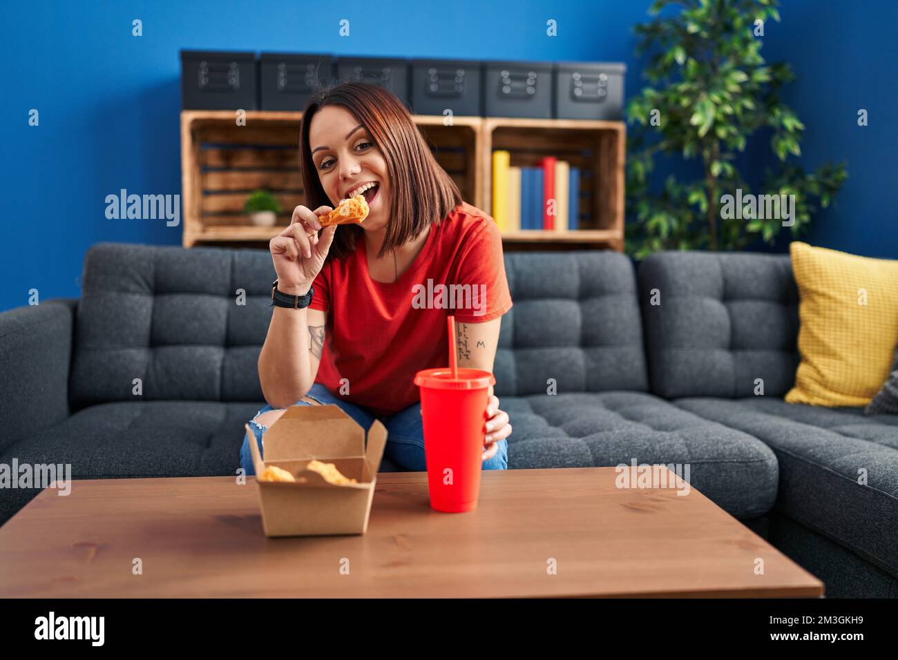 Young beautiful hispanic woman eating fried chicken sitting on sofa at ...