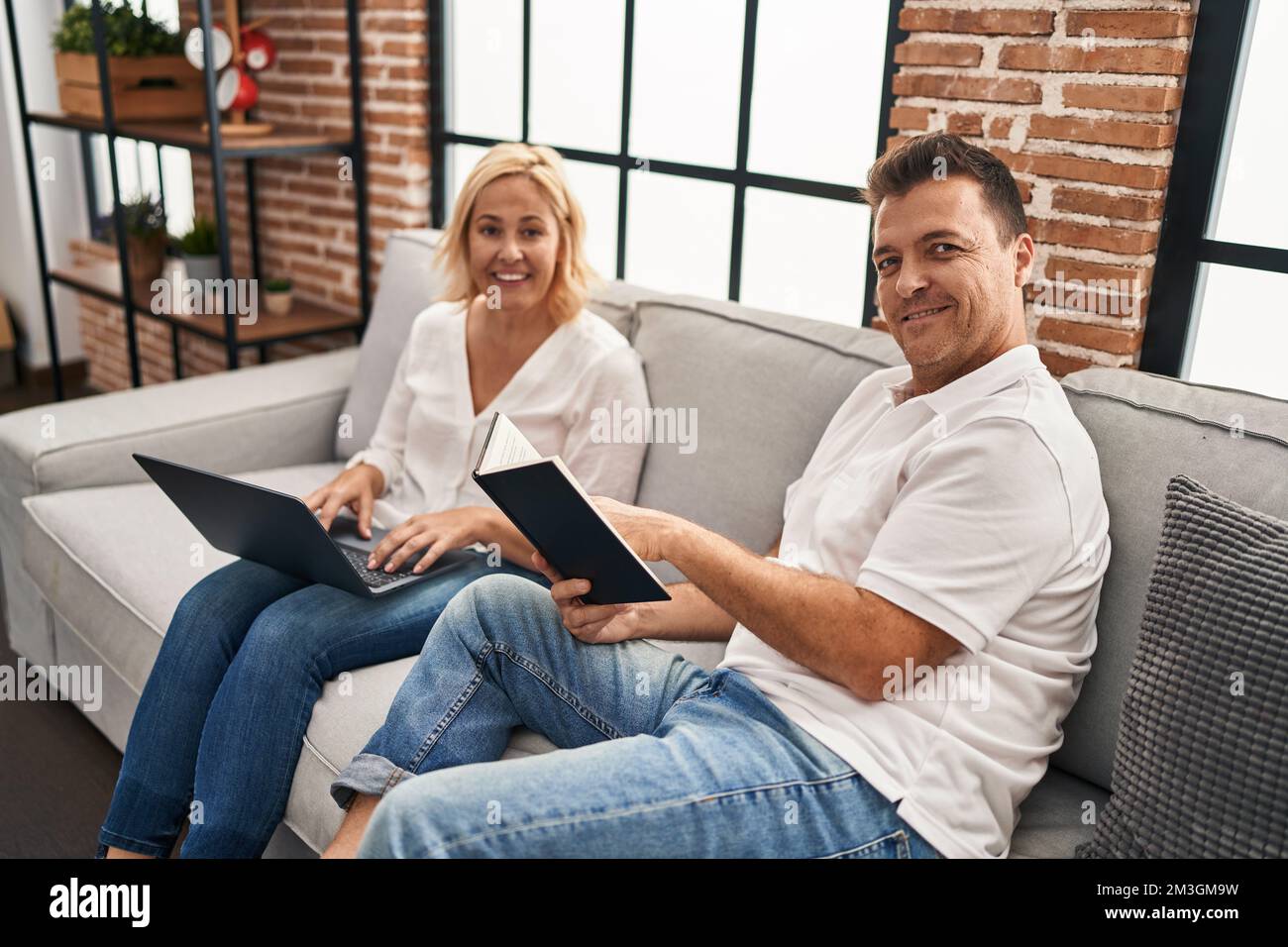 Middle age man and woman using laptop and reading book sitting on sofa ...