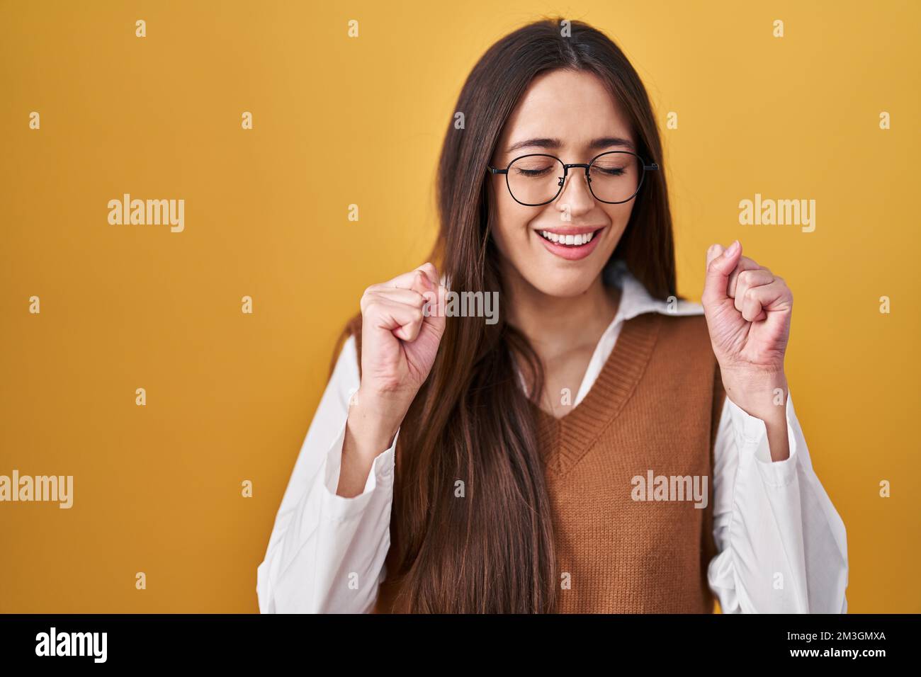 Young brunette woman standing over yellow background wearing glasses ...