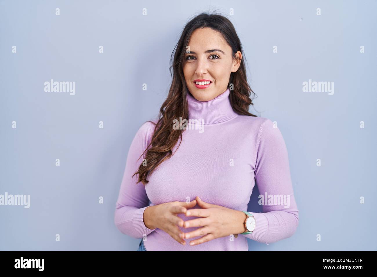 Young brunette woman standing over blue background hands together and ...