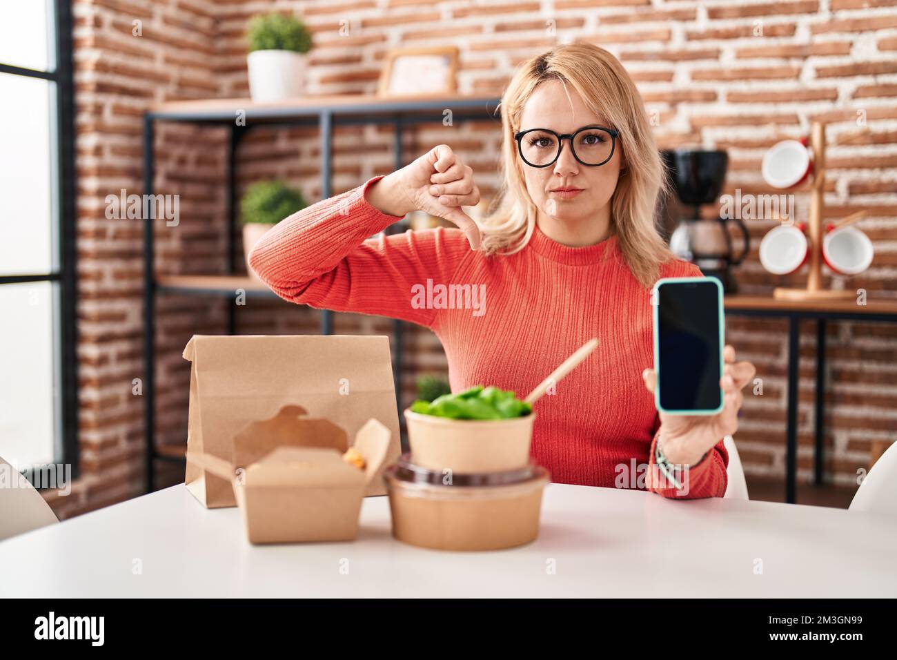 Blonde woman eating take away food showing smartphone screen with angry ...