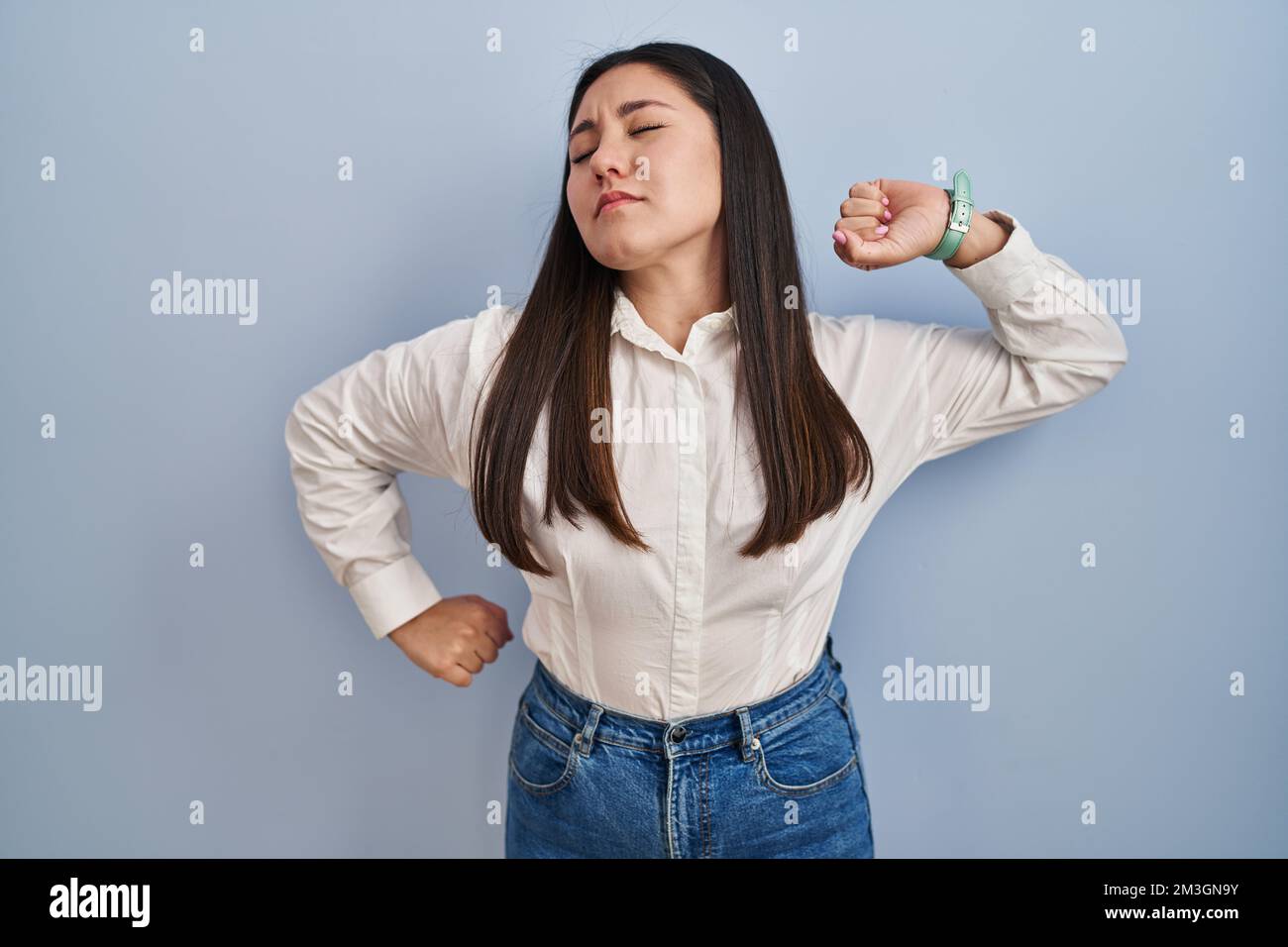 Young latin woman standing over blue background stretching back, tired ...