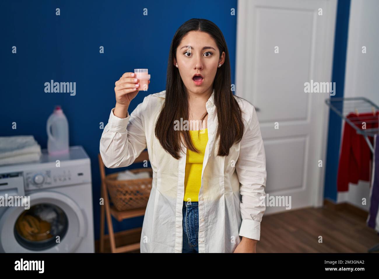 Young latin woman doing laundry holding detergent scared and amazed with open mouth for surprise ...