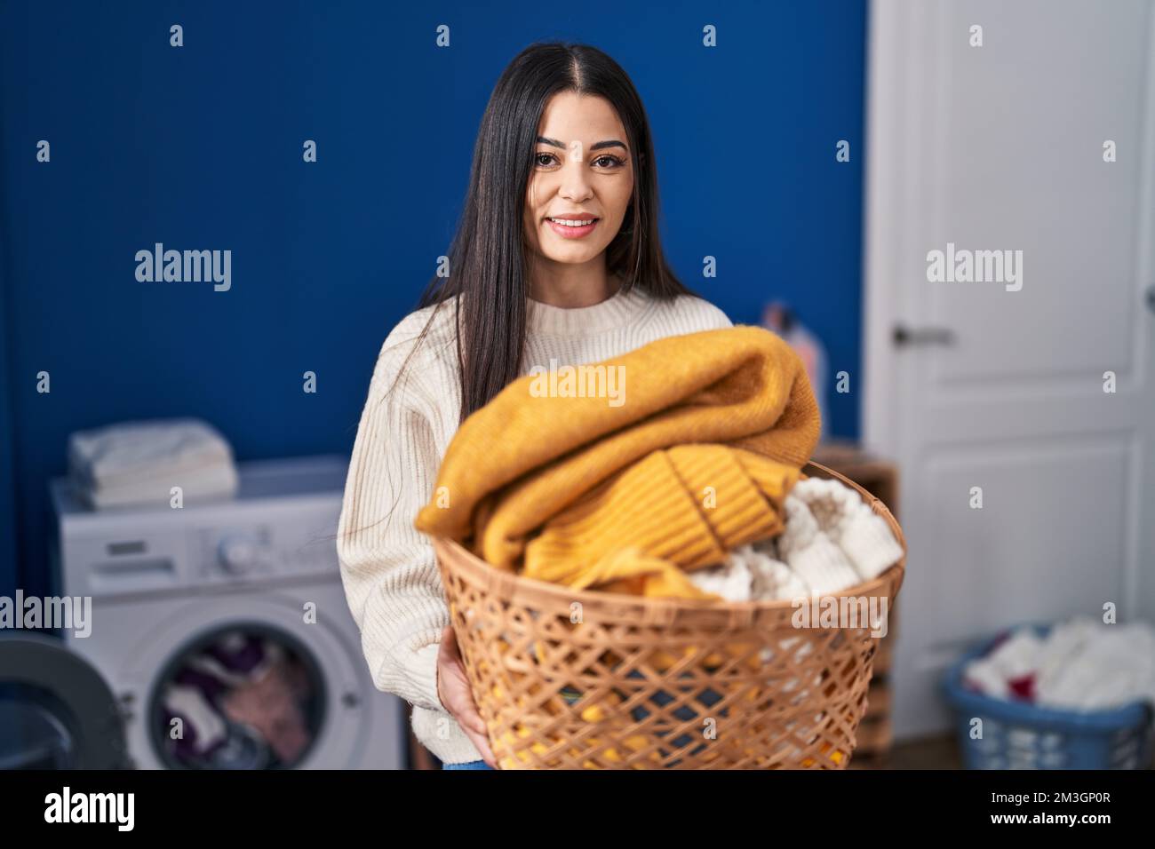 Young woman holding laundry basket smiling with a happy and cool smile ...