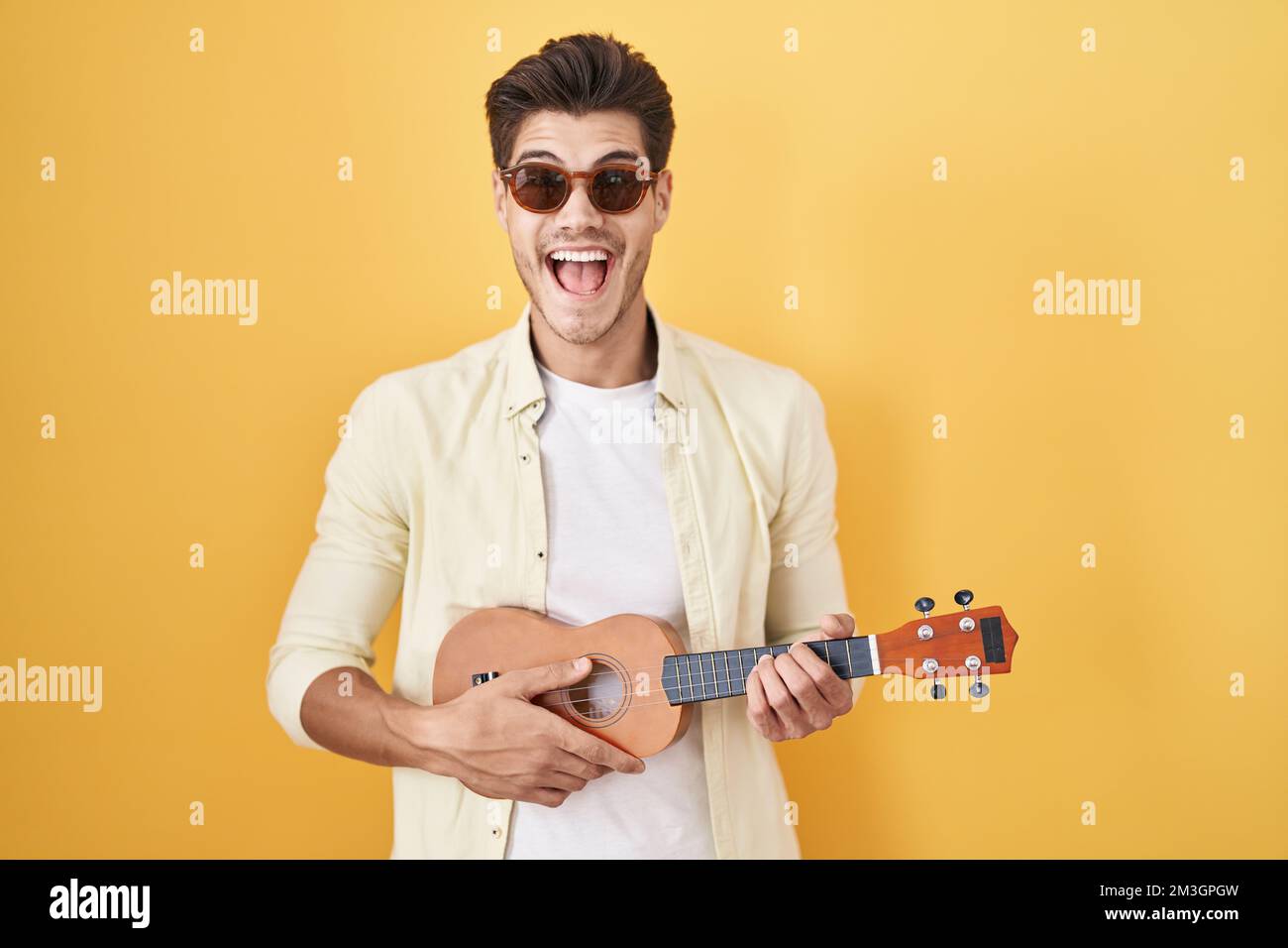 Young hispanic man playing ukulele over yellow background celebrating ...