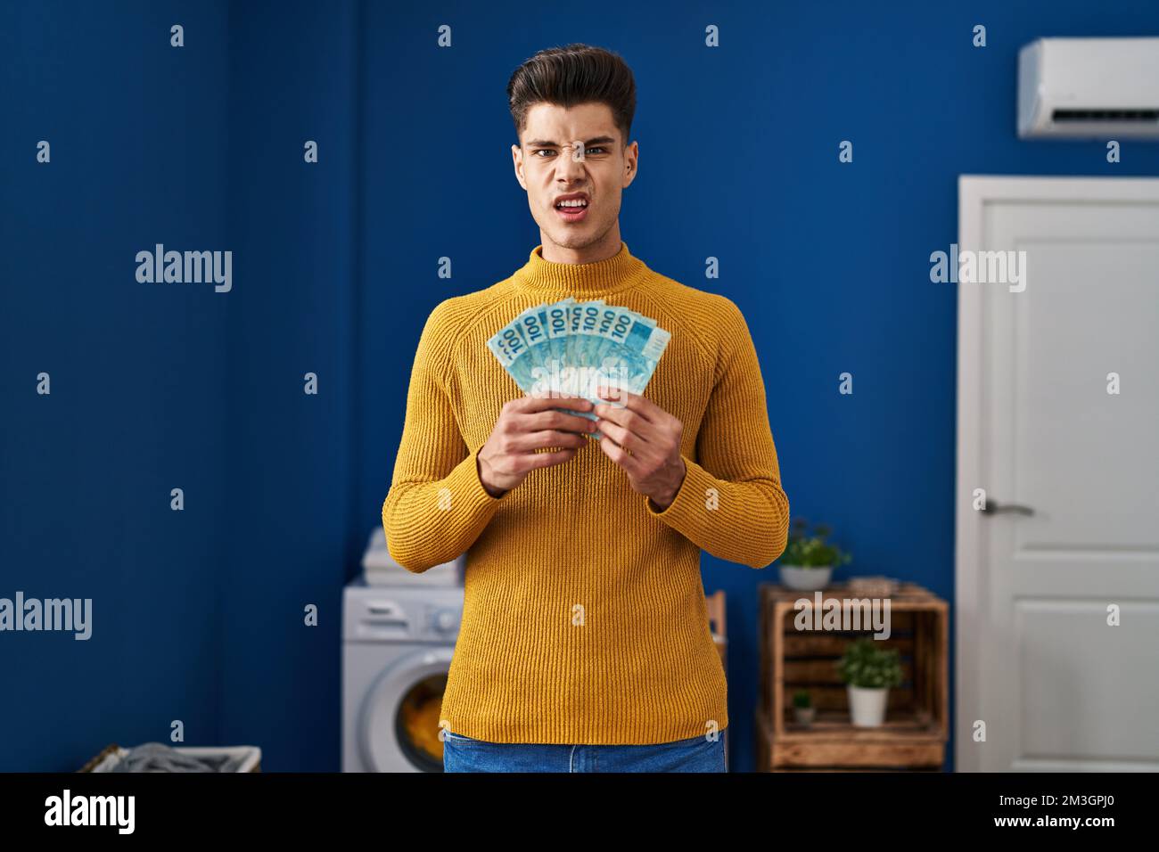 Young hispanic man at laundry room holding brazilian reals clueless and ...