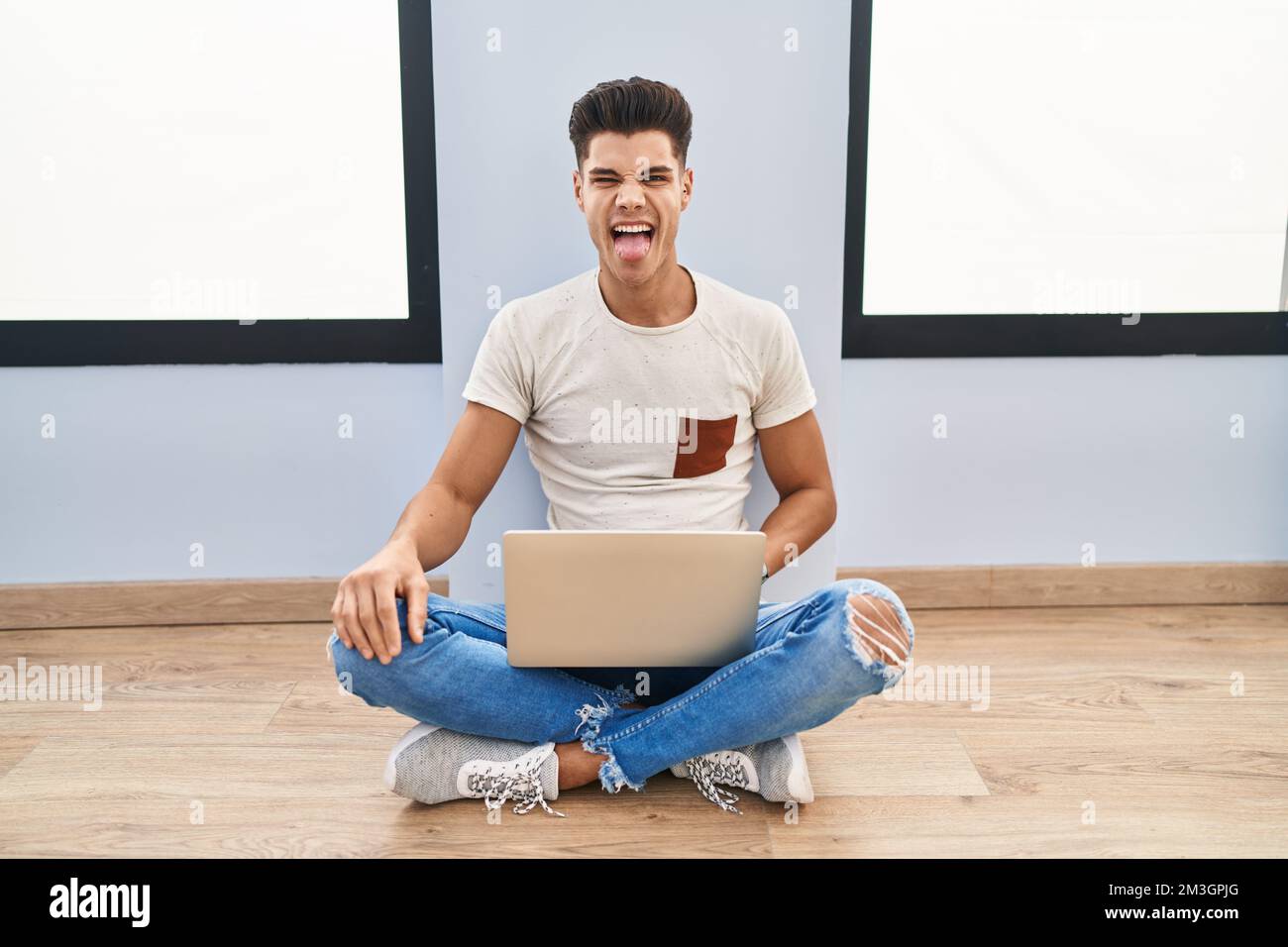Young hispanic man using laptop at home sticking tongue out happy with ...