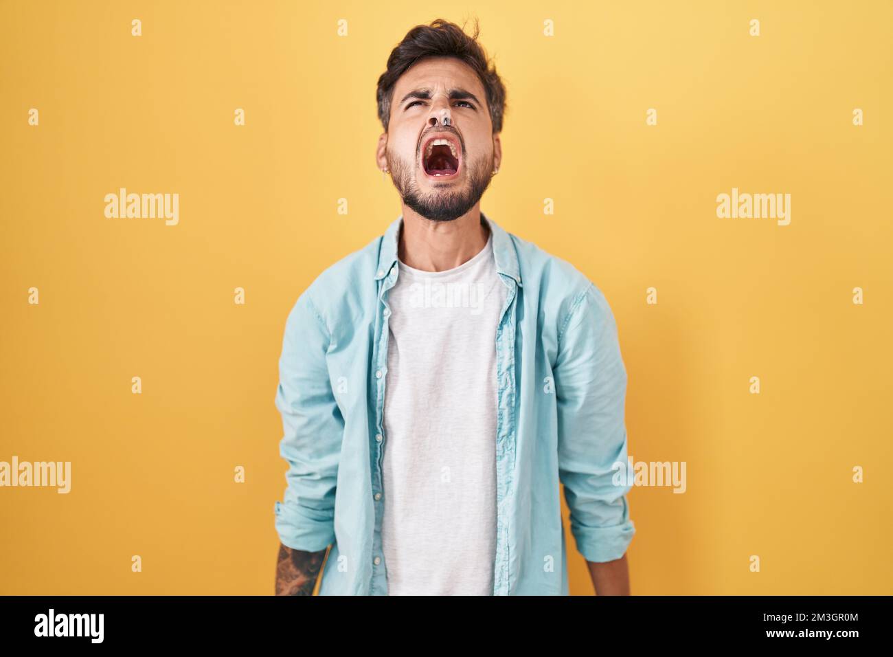 Young hispanic man with tattoos standing over yellow background angry ...
