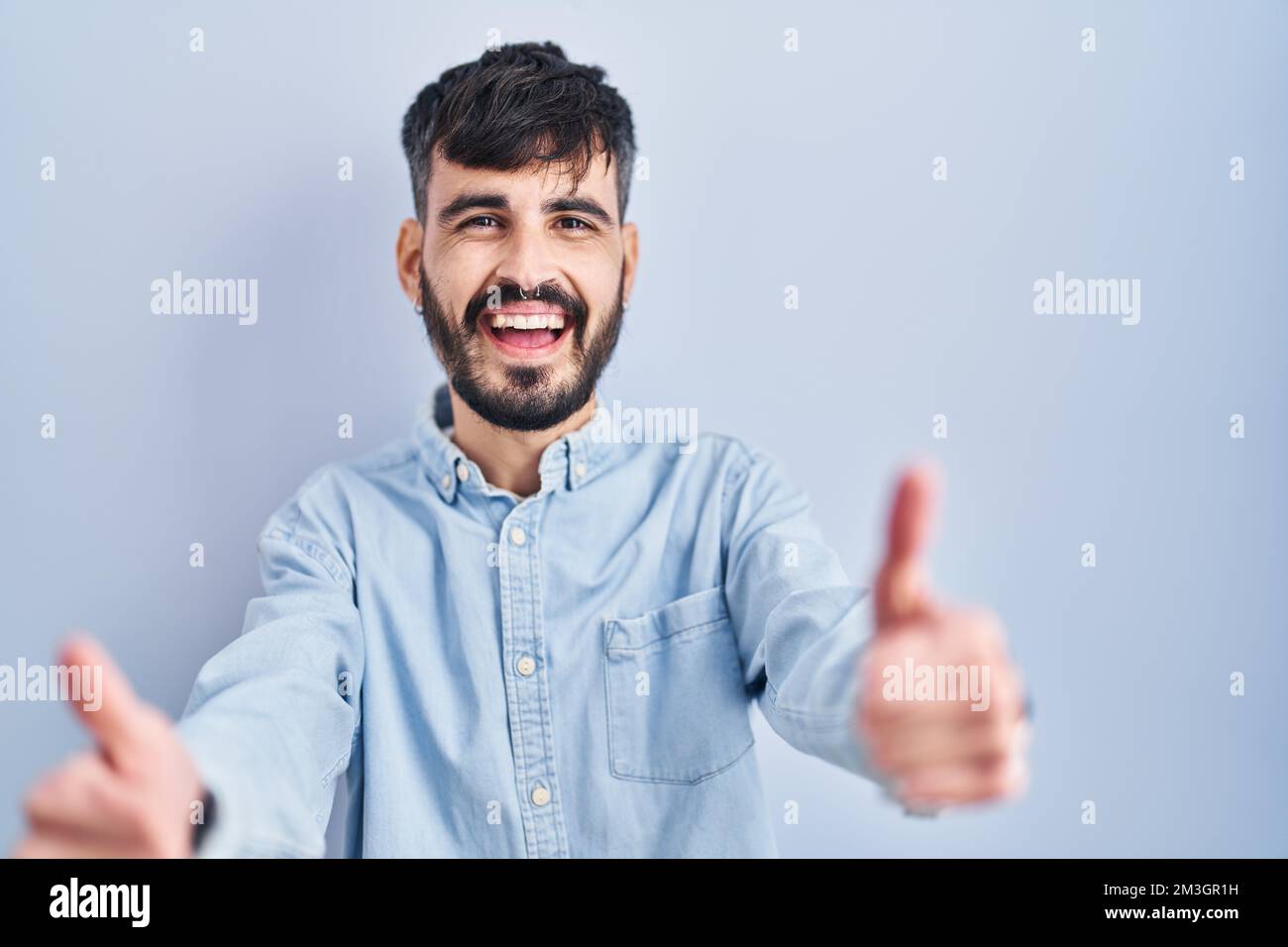 Young hispanic man with beard standing over blue background approving ...