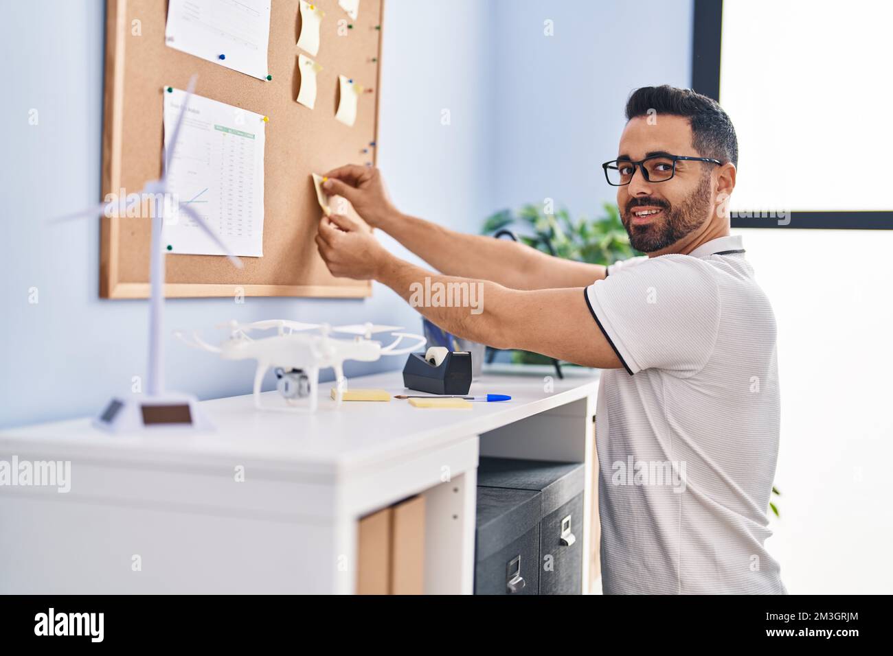 Young hispanic man business worker hanging reminder paper on cork board ...