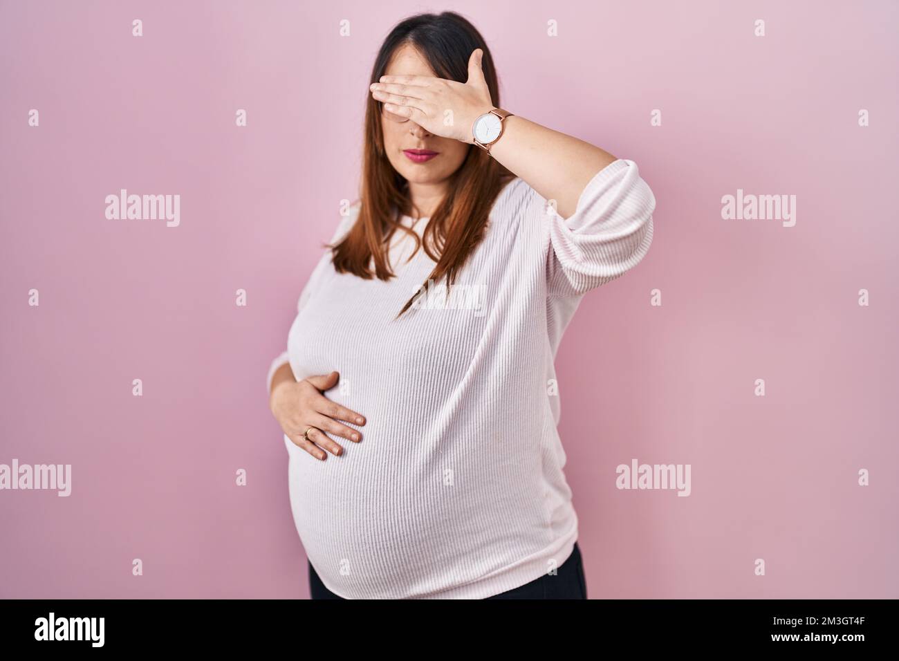 Pregnant woman standing over pink background covering eyes with hand ...