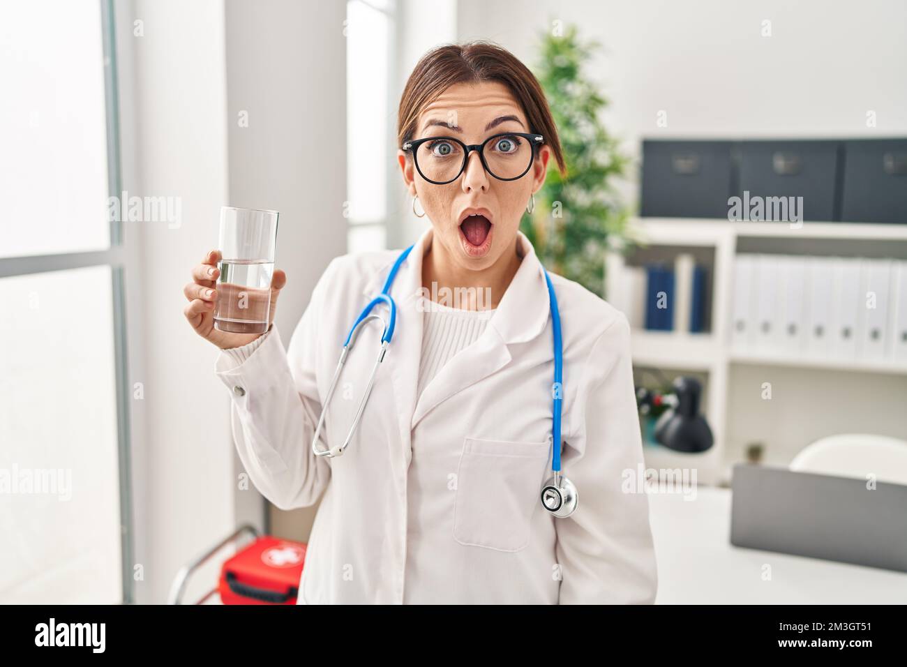 Young brunette doctor woman holding glass of water scared and amazed ...