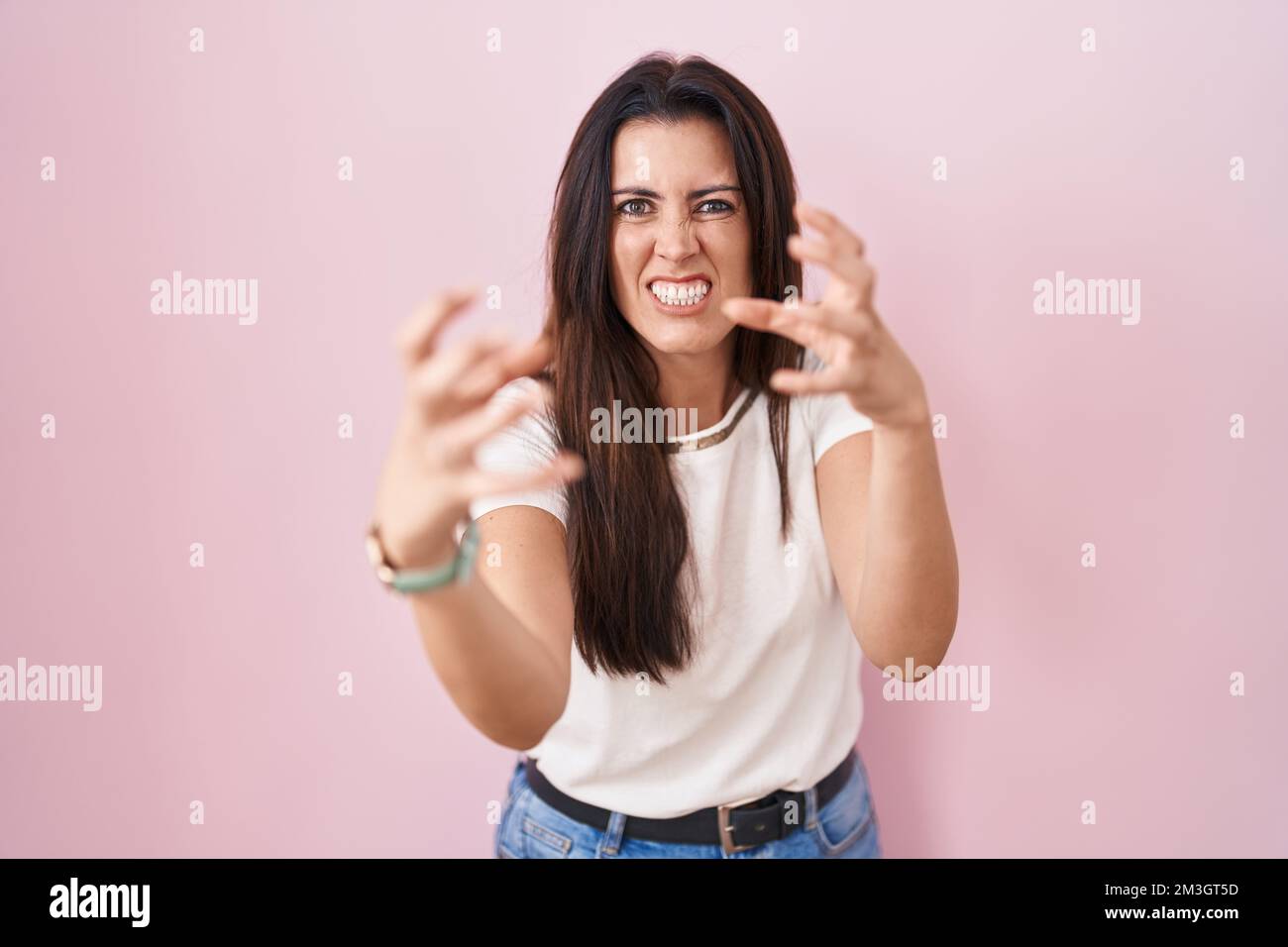 Young brunette woman standing over pink background shouting frustrated ...