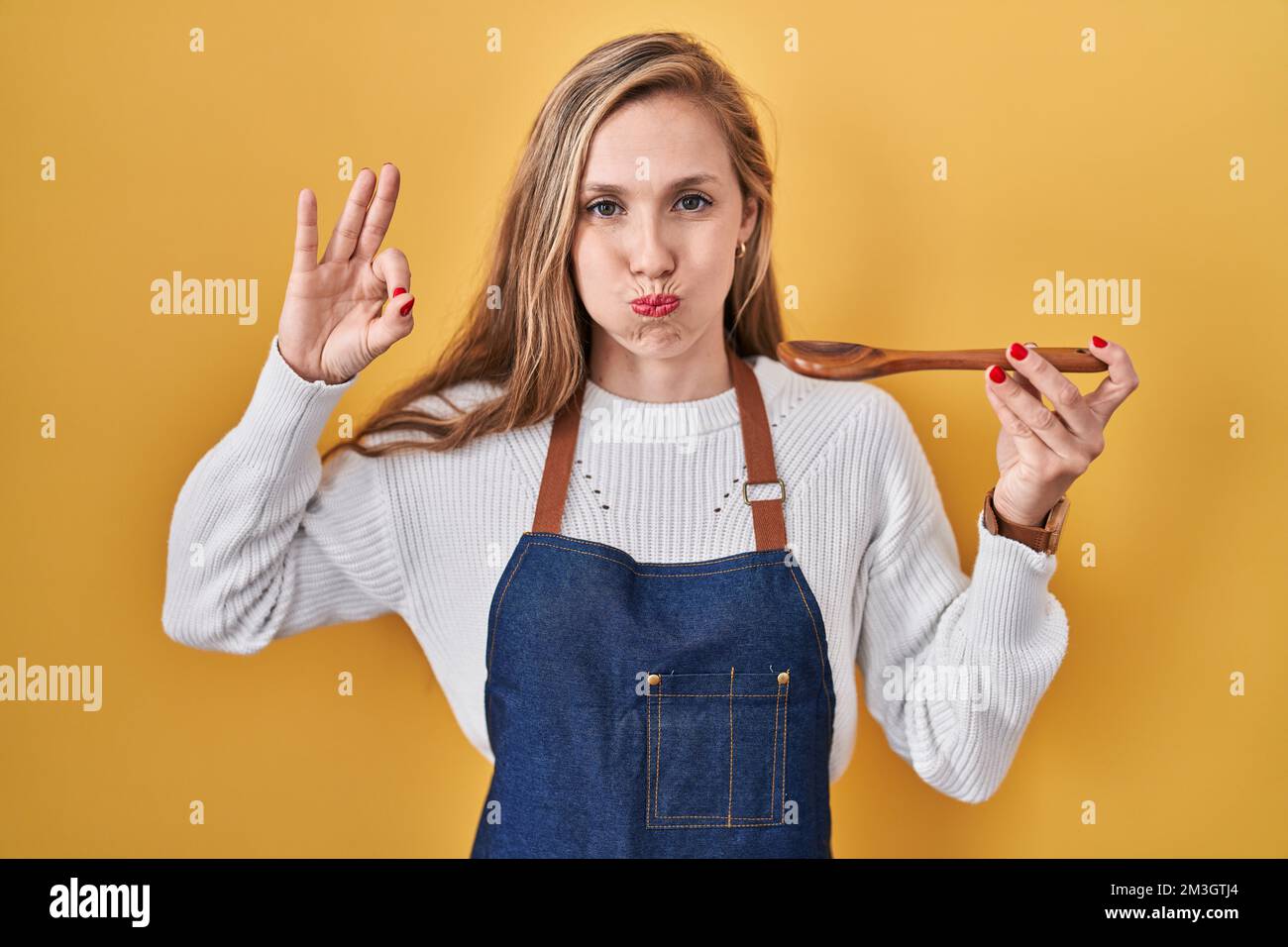 Young blonde woman wearing apron tasting food holding wooden spoon ...