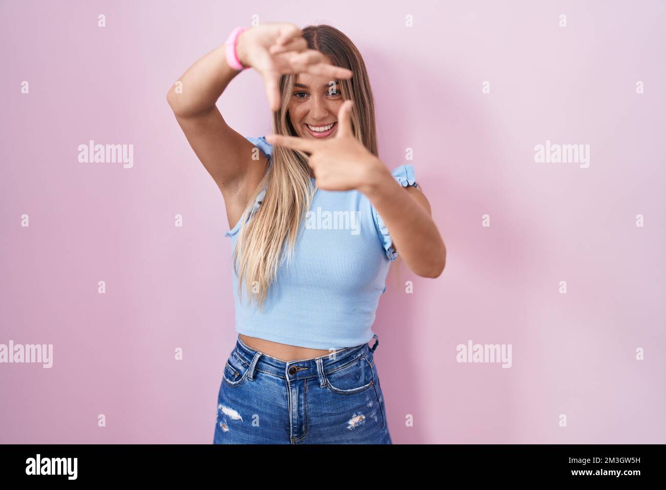 Young blonde woman standing over pink background smiling making frame ...