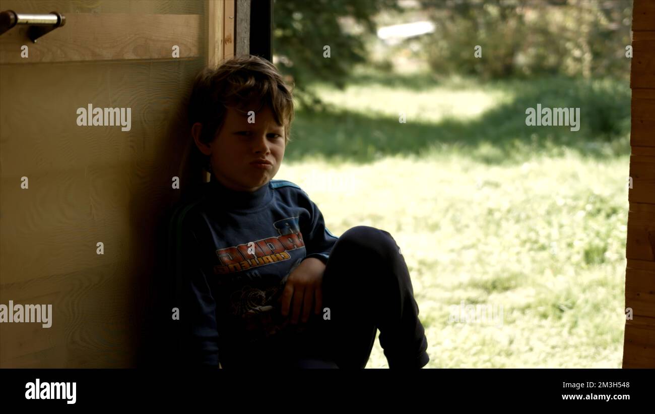 Boy child looking sad sitting by the opened door of a house with summer ...