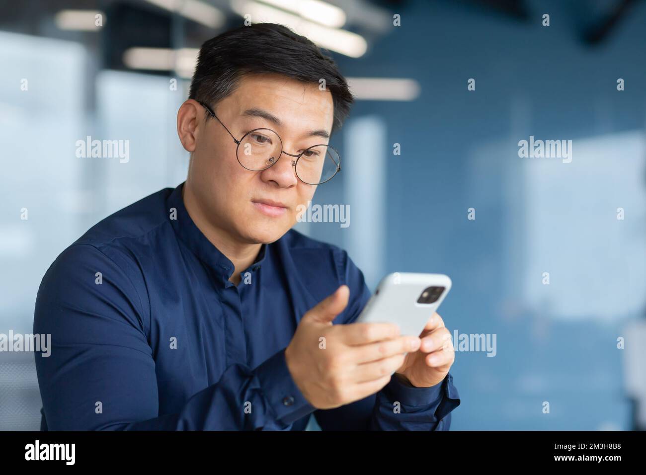 Close-up of serious and pensive Asian businessman using phone, man inside office typing message ...