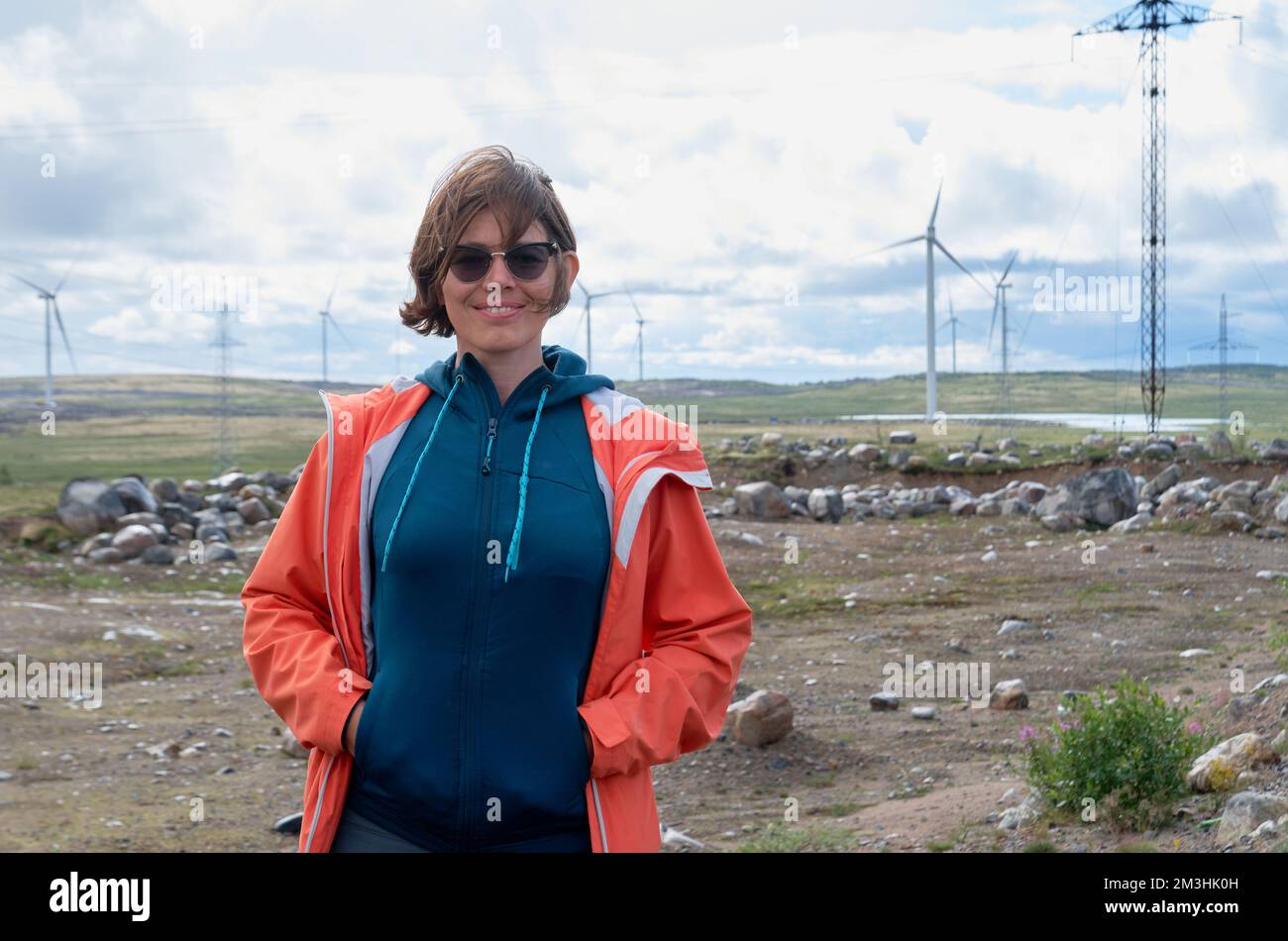 Smiling woman in orange jacket and sunglasses in front of wind farm ...