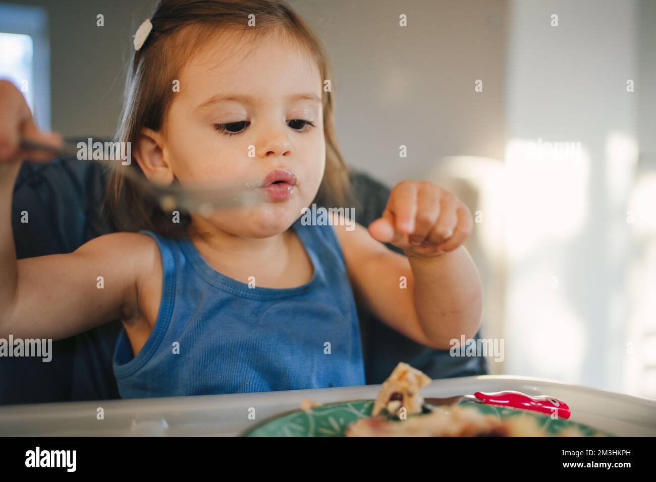 Messy baby girl eating with a spoon in high chair. Daily routine. Finger food. Healthy child ...