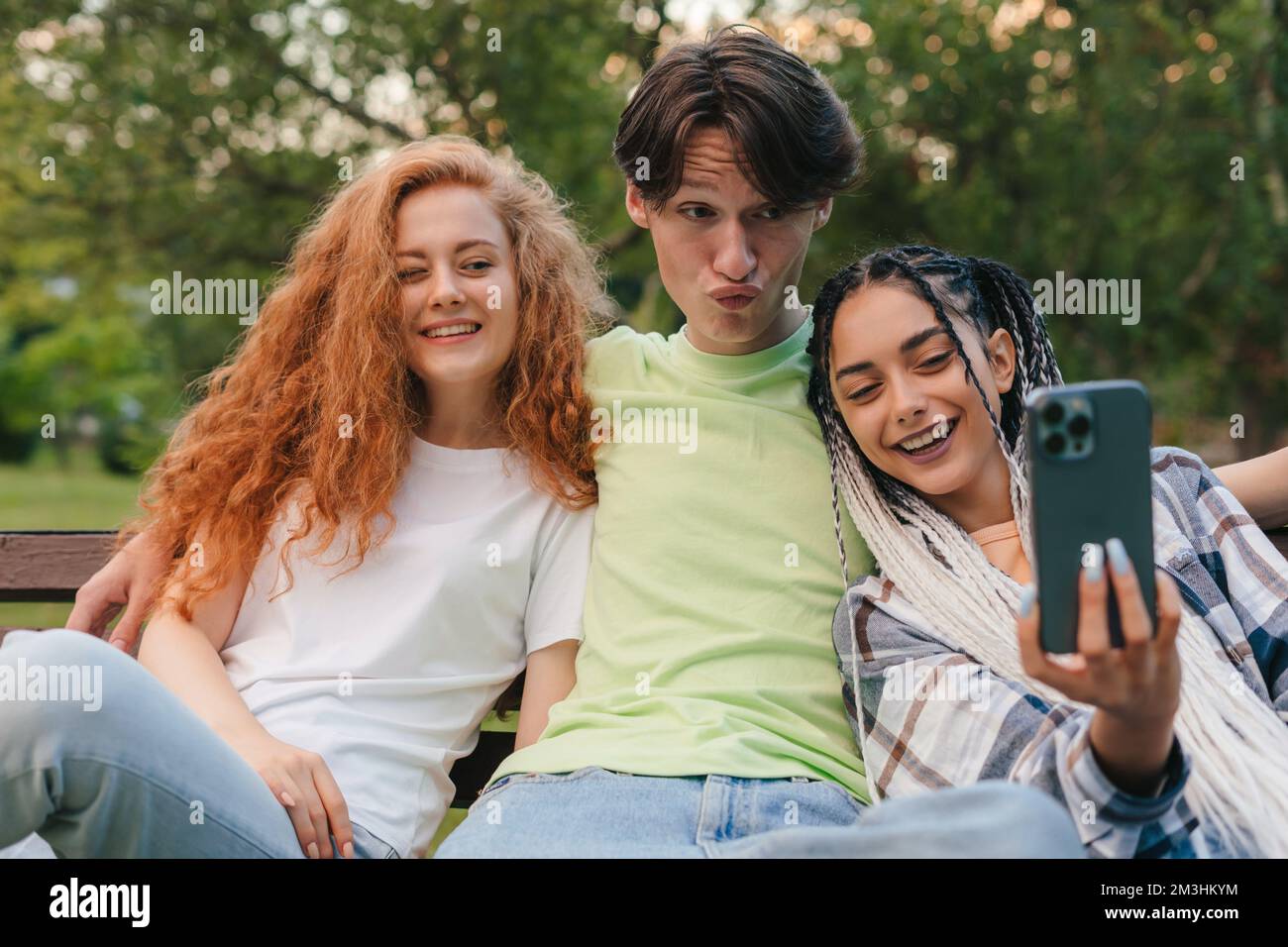 Cheerful smiling friends at the park sitting on a bench and taking ...