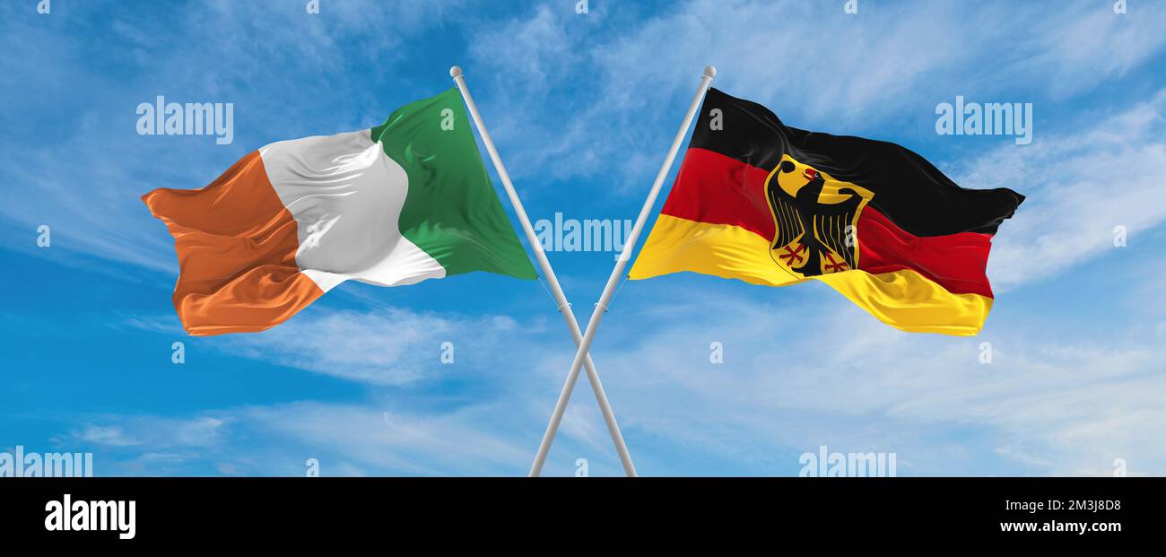 two crossed flags Germany and Ireland waving in wind at cloudy sky ...