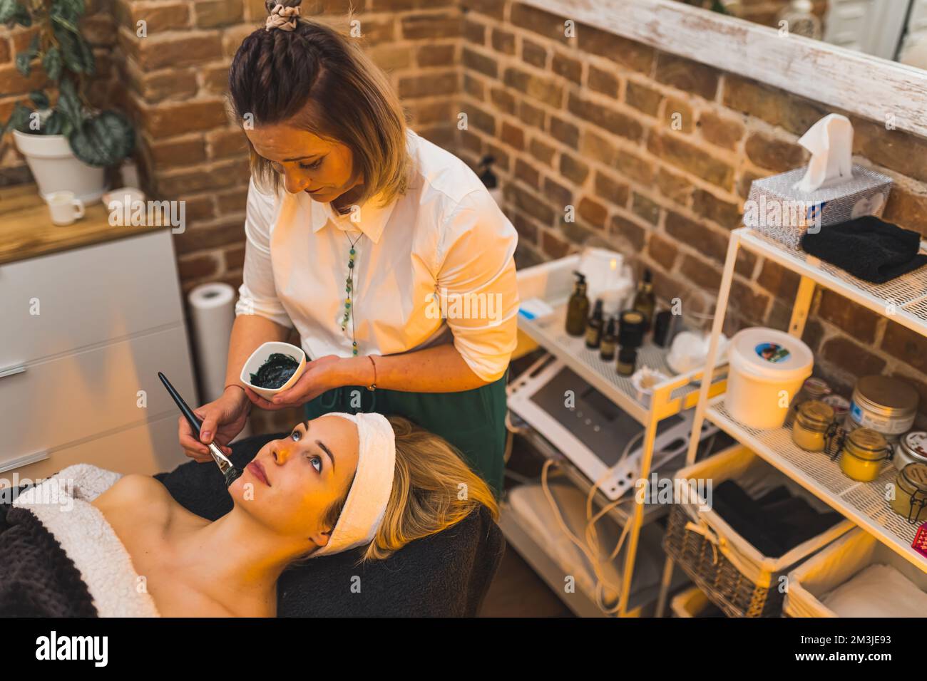 Cosmetologist applying black mask on the face of a beautiful blond ...