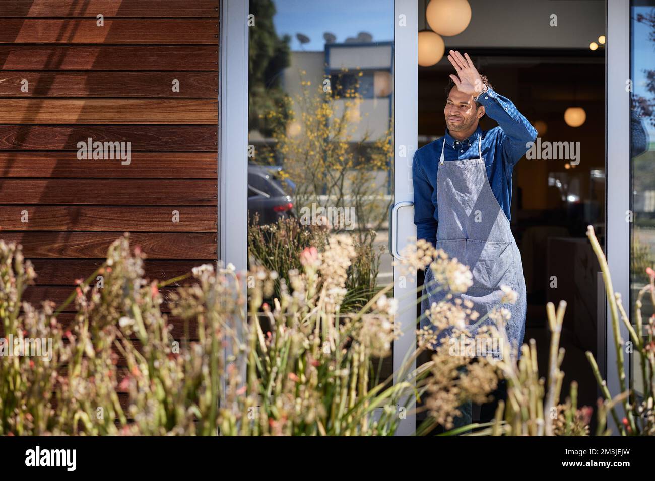 Smiling male entrepreneur waving while leaning on door at coffee shop ...