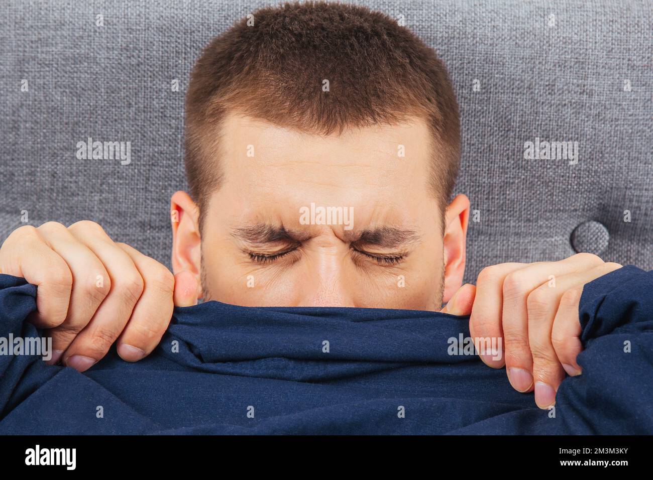 With closed eyes. A handsome young guy hiding behind a blanket looks to ...