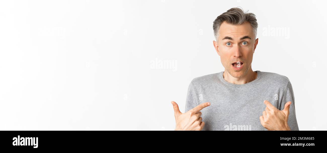 Close-up of confused middle-aged man, pointing at himself and gasping, standing over white ...